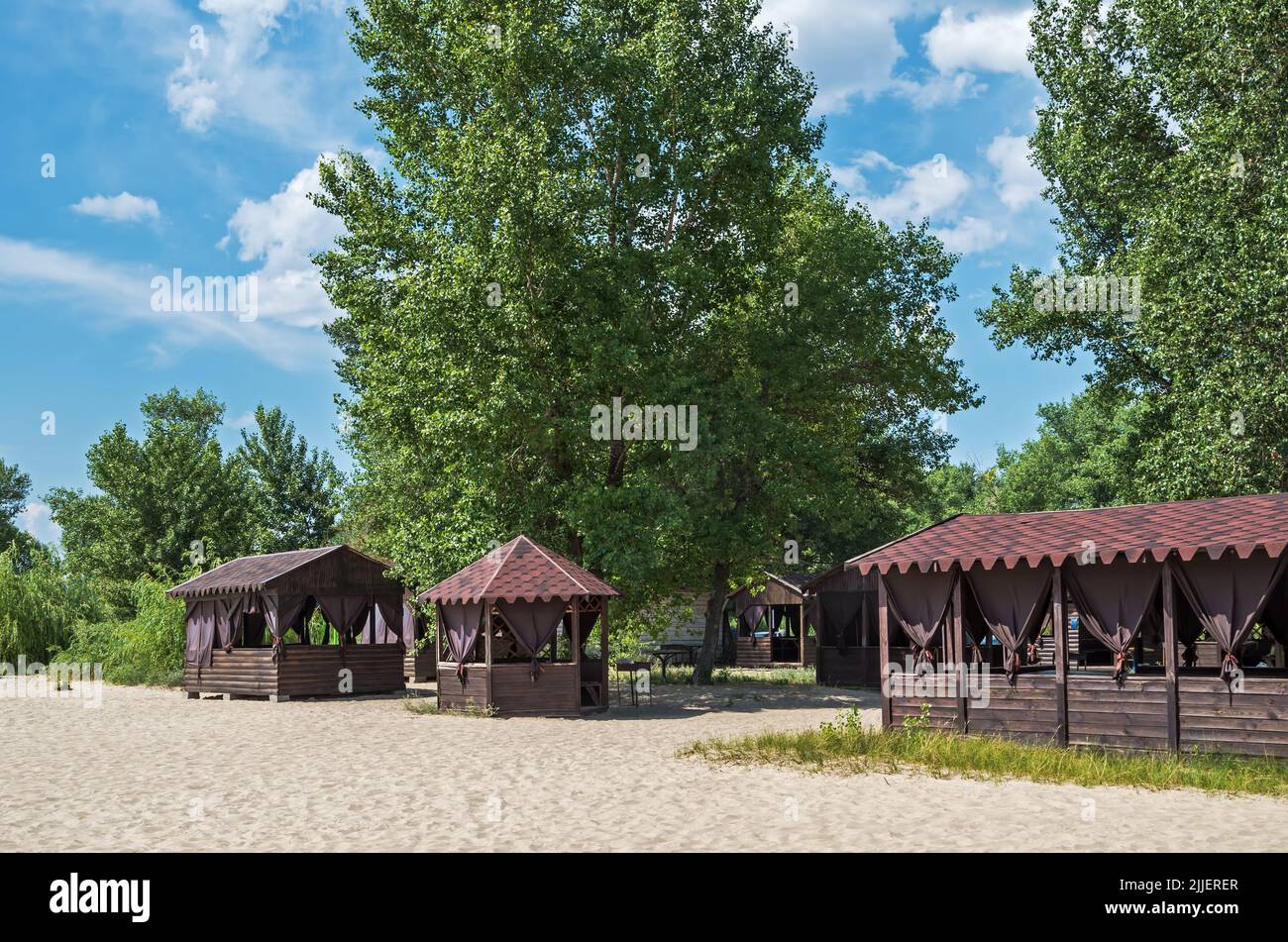 Wooden bowers and tents for relaxing on river beach in shade of branchy ...