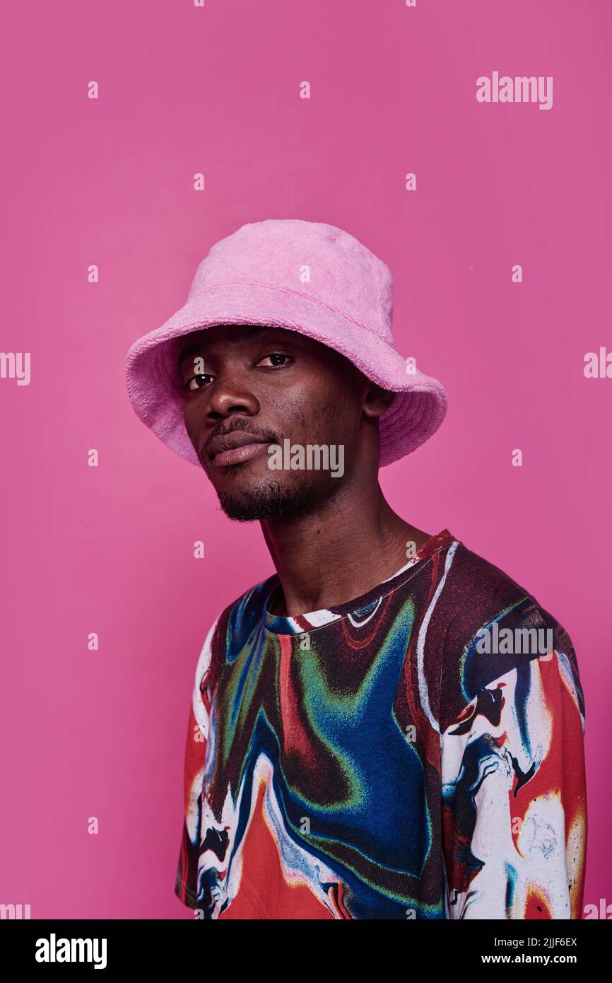 Portrait of African teenage boy in panama hat and bright shirt posing against pink background ...
