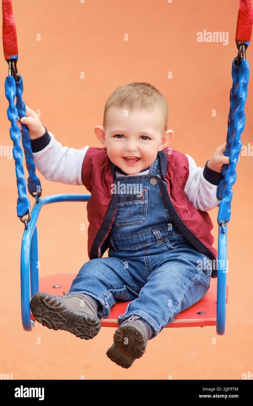 Happy toddler baby boy rides on a swing. Smiling child swinging on the playground, kid aged one ...