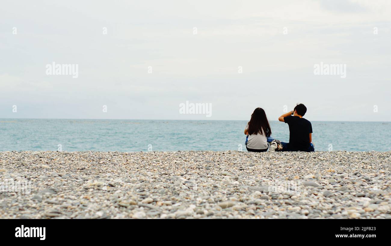 A back view of young couple sitting in rocky beach in background of sea ...