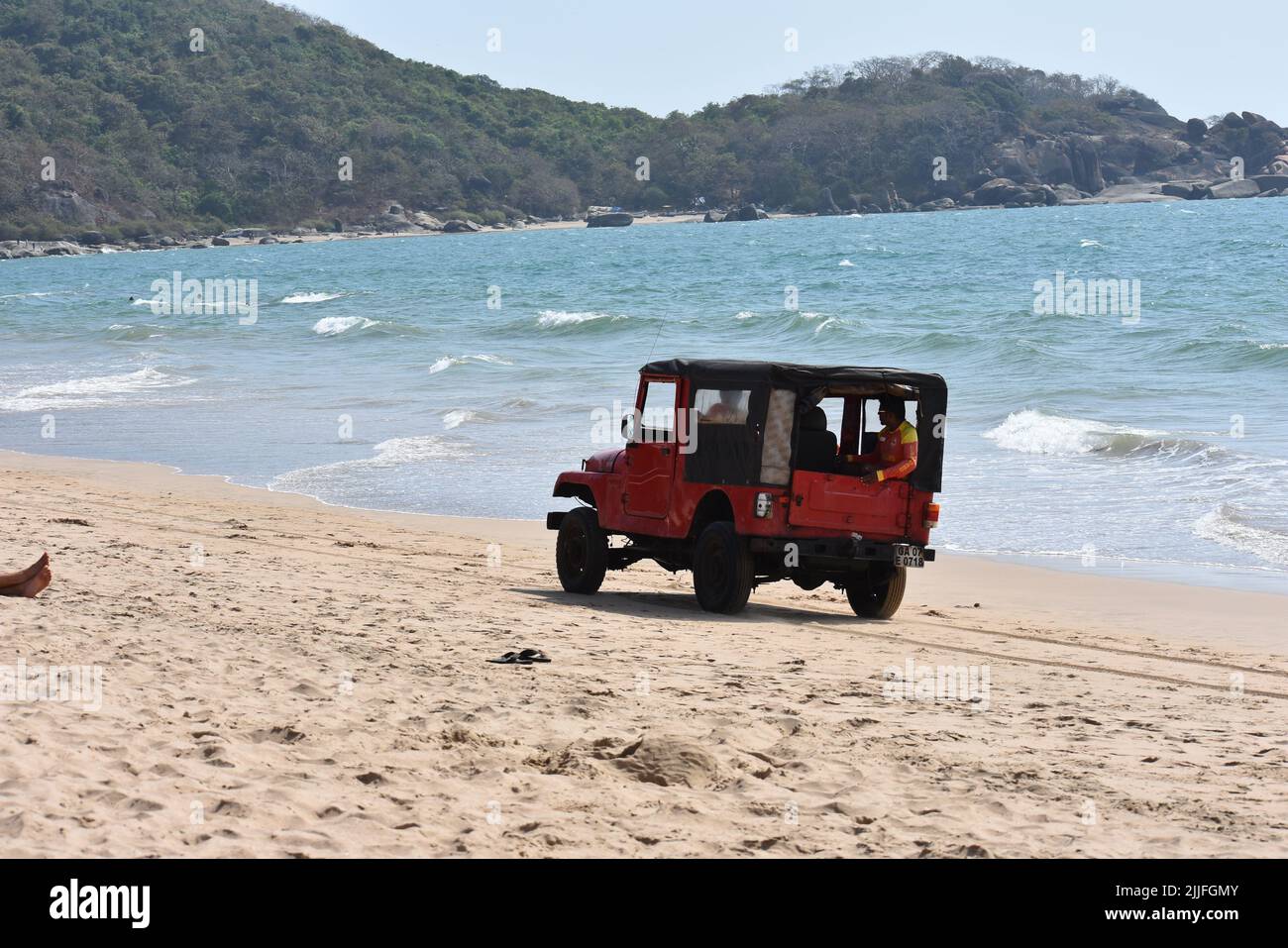 A red jeep car driving on a beach near a wavy sea with a hill in the ...