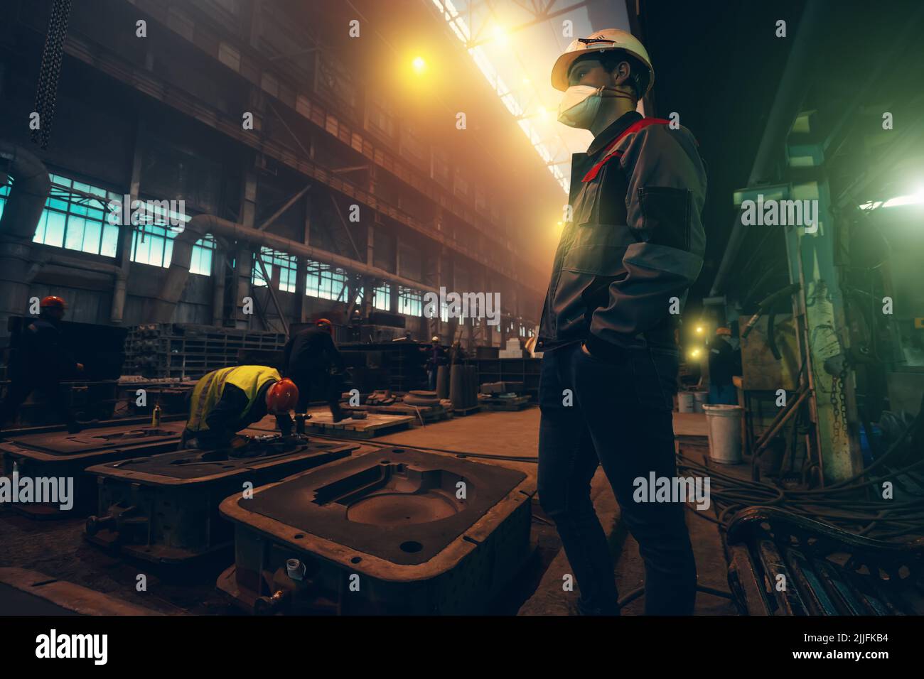 Engineer in helmet, face mask and uniform controls workers in workshop ...