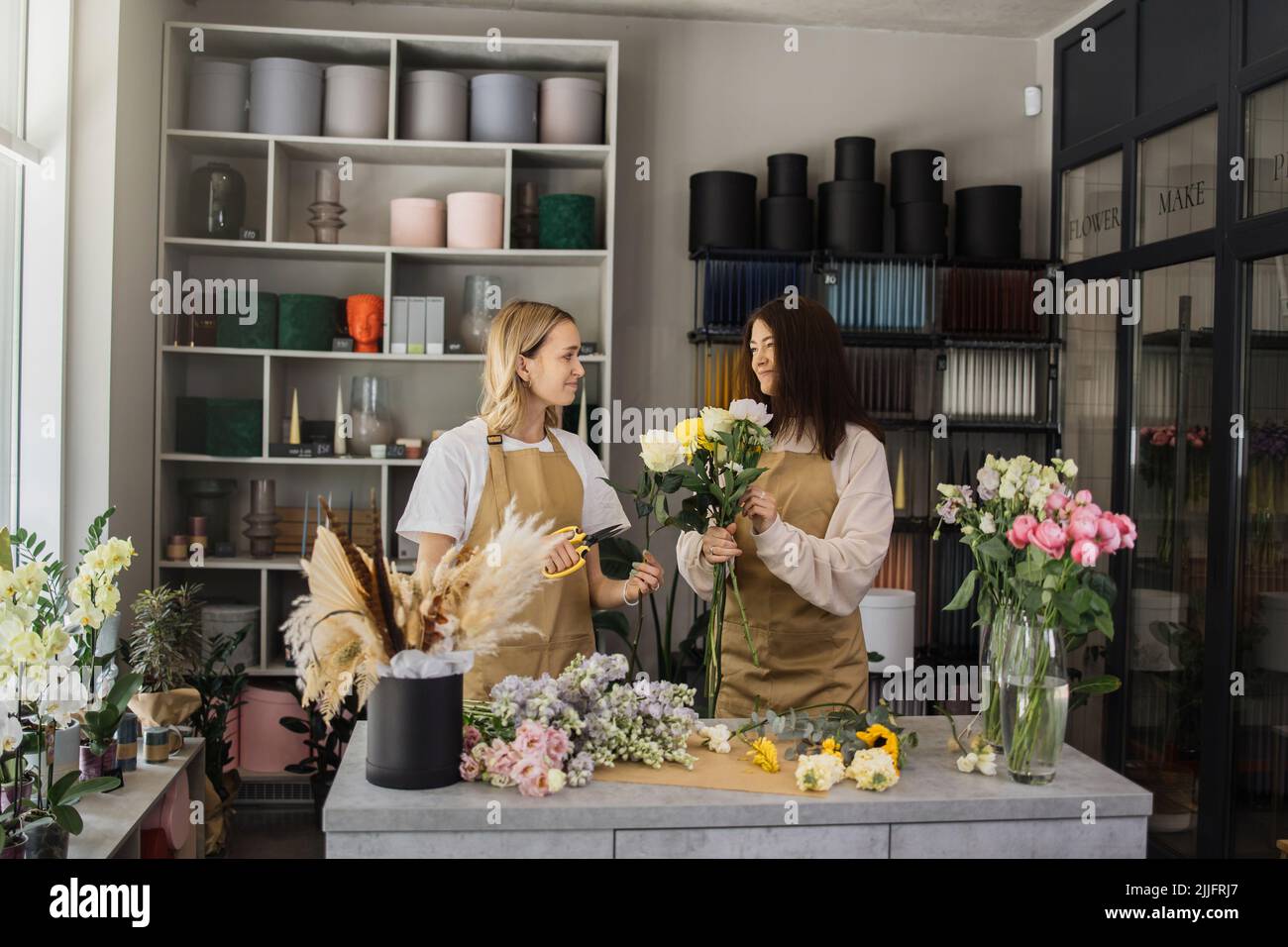 Two smiling florists in uniform work in a flower shop behind counter with different varieties of ...