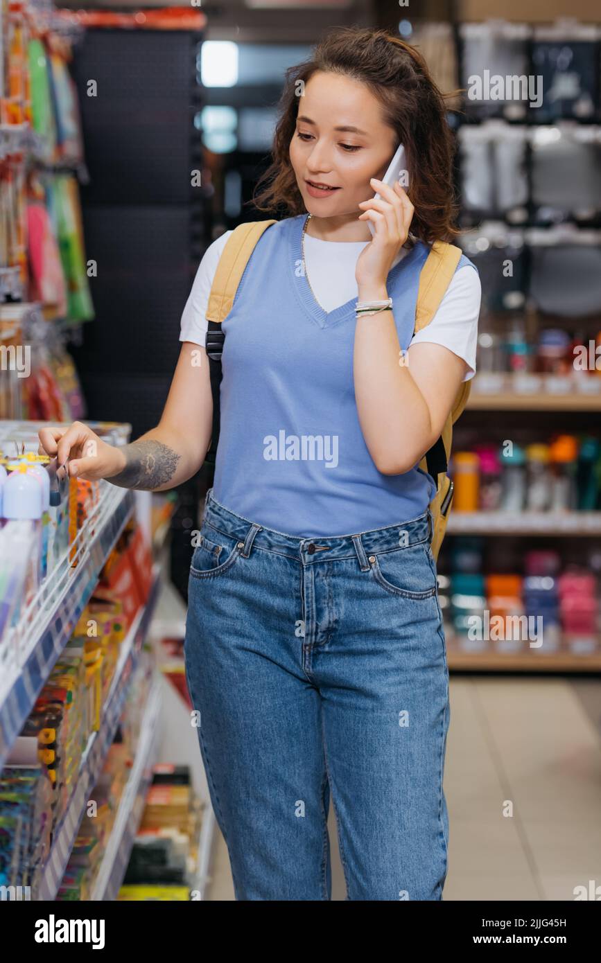 tattooed woman talking on mobile phone near rack in stationery shop ...