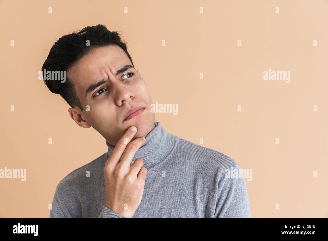 Young puzzled man frowning while holding his chin isolated over beige ...