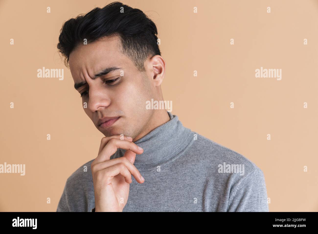 Young puzzled man frowning while holding his chin isolated over beige ...