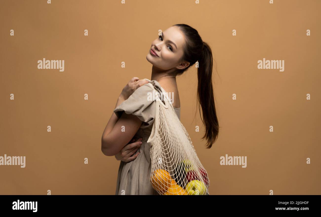 Young brunette woman with cotton reusable mesh bag full of fruits ...