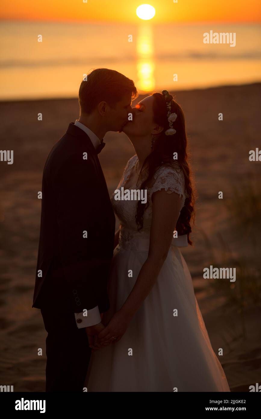 The vertical view of the bride and groom kissing on the shore at sunset ...