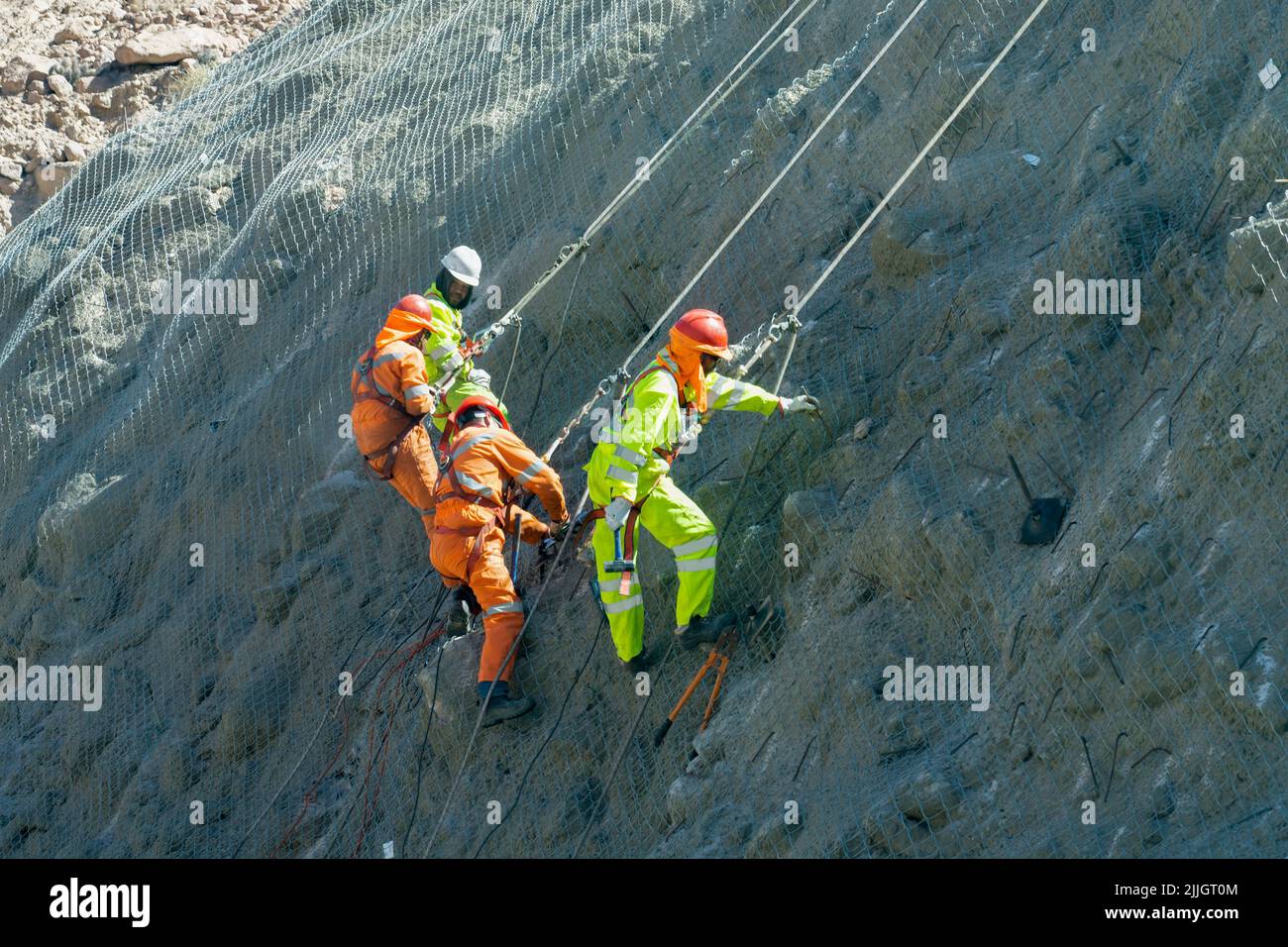 Construction workers on ropes set rebar anchors on wire netting to ...