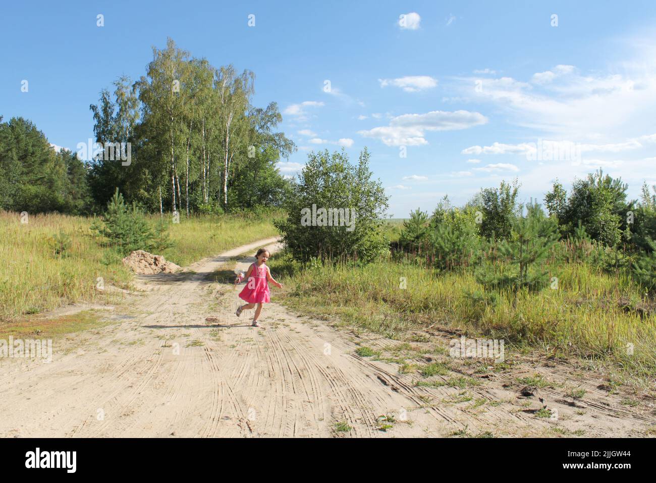 Funny toddler girl in pink dress with a doll runs along sandy path ...