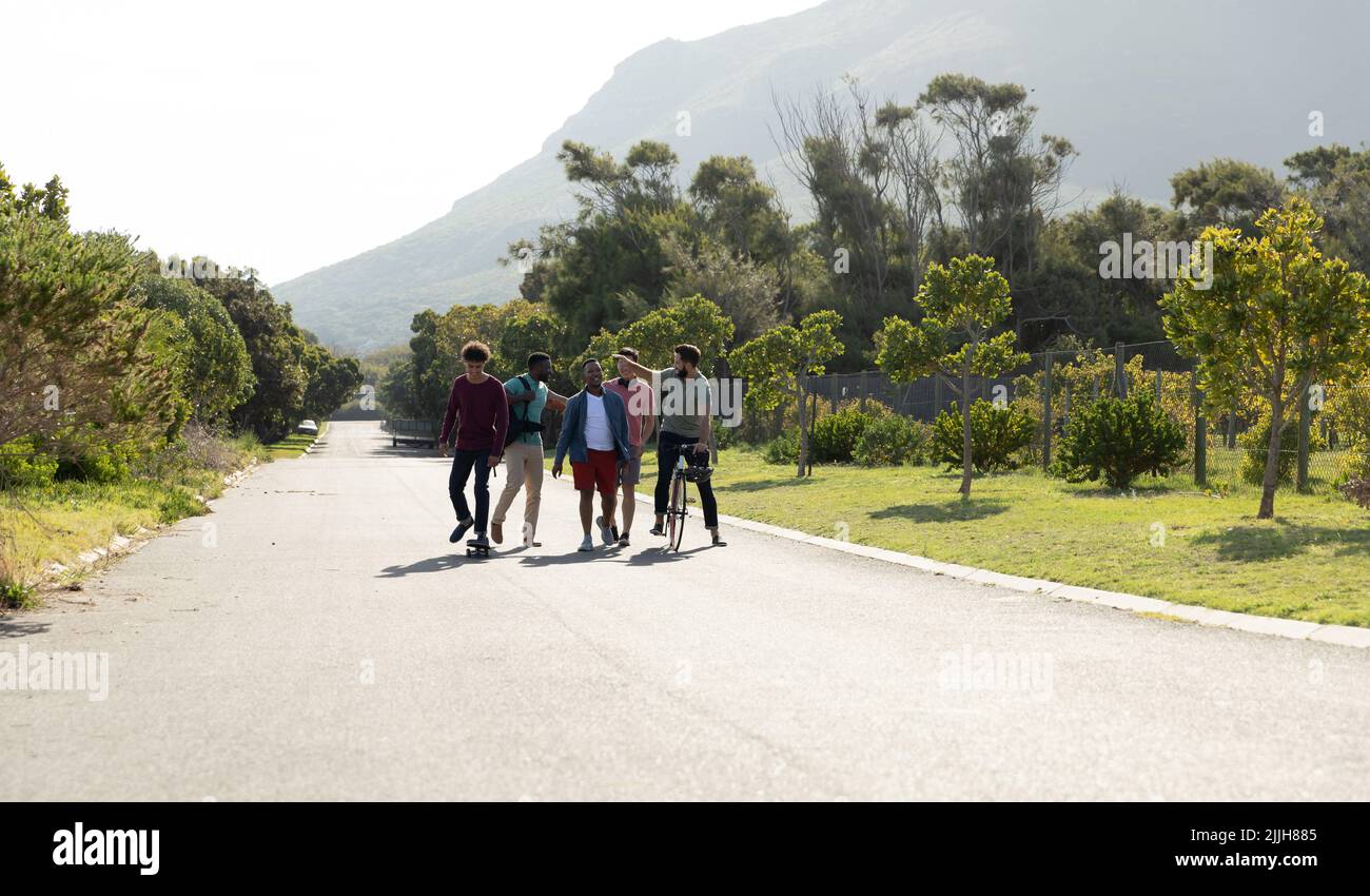 Male multiracial friends with bicycle walking on road amidst lush trees ...