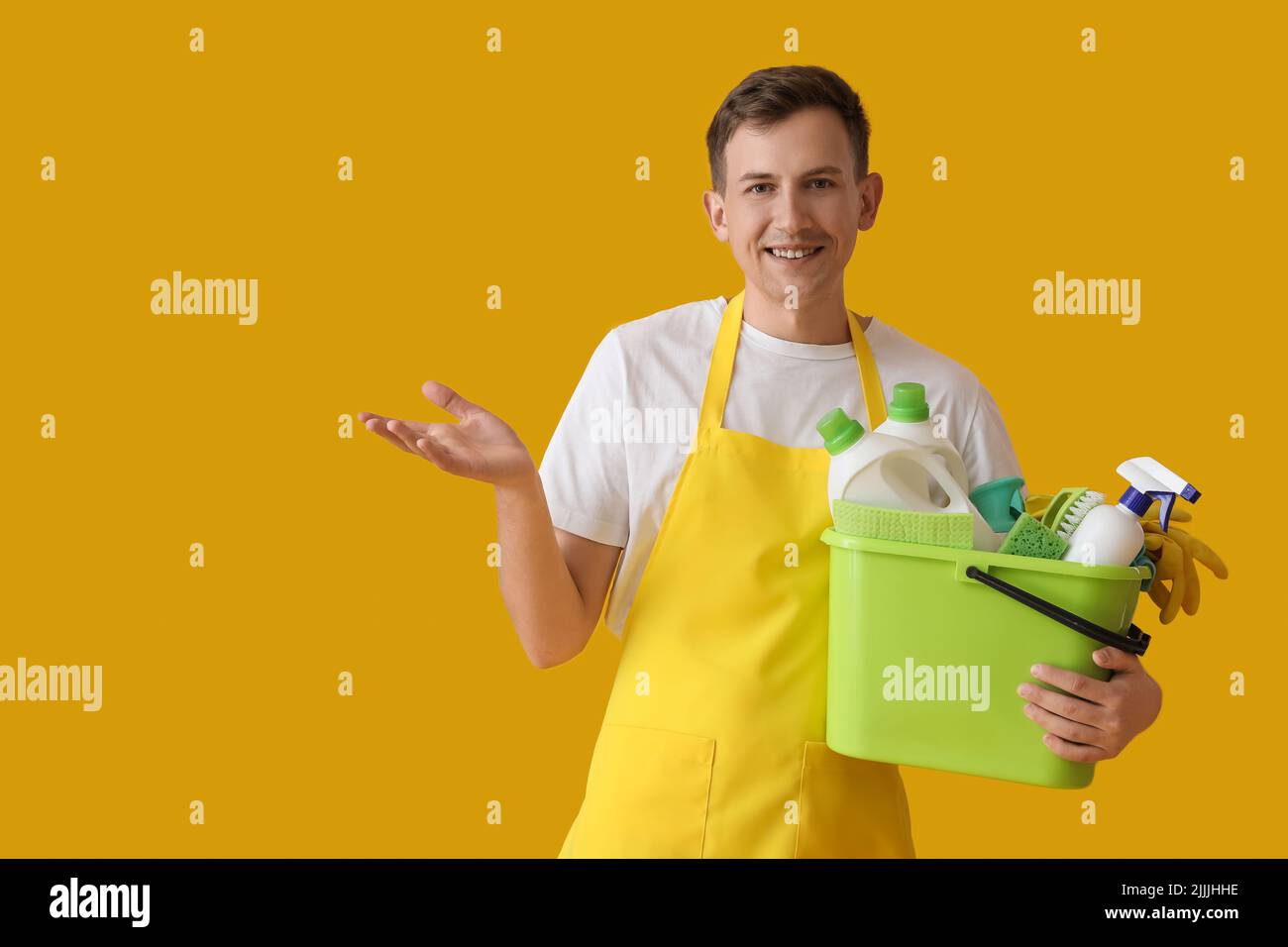 Young man holding bucket with cleaning supplies on yellow background ...