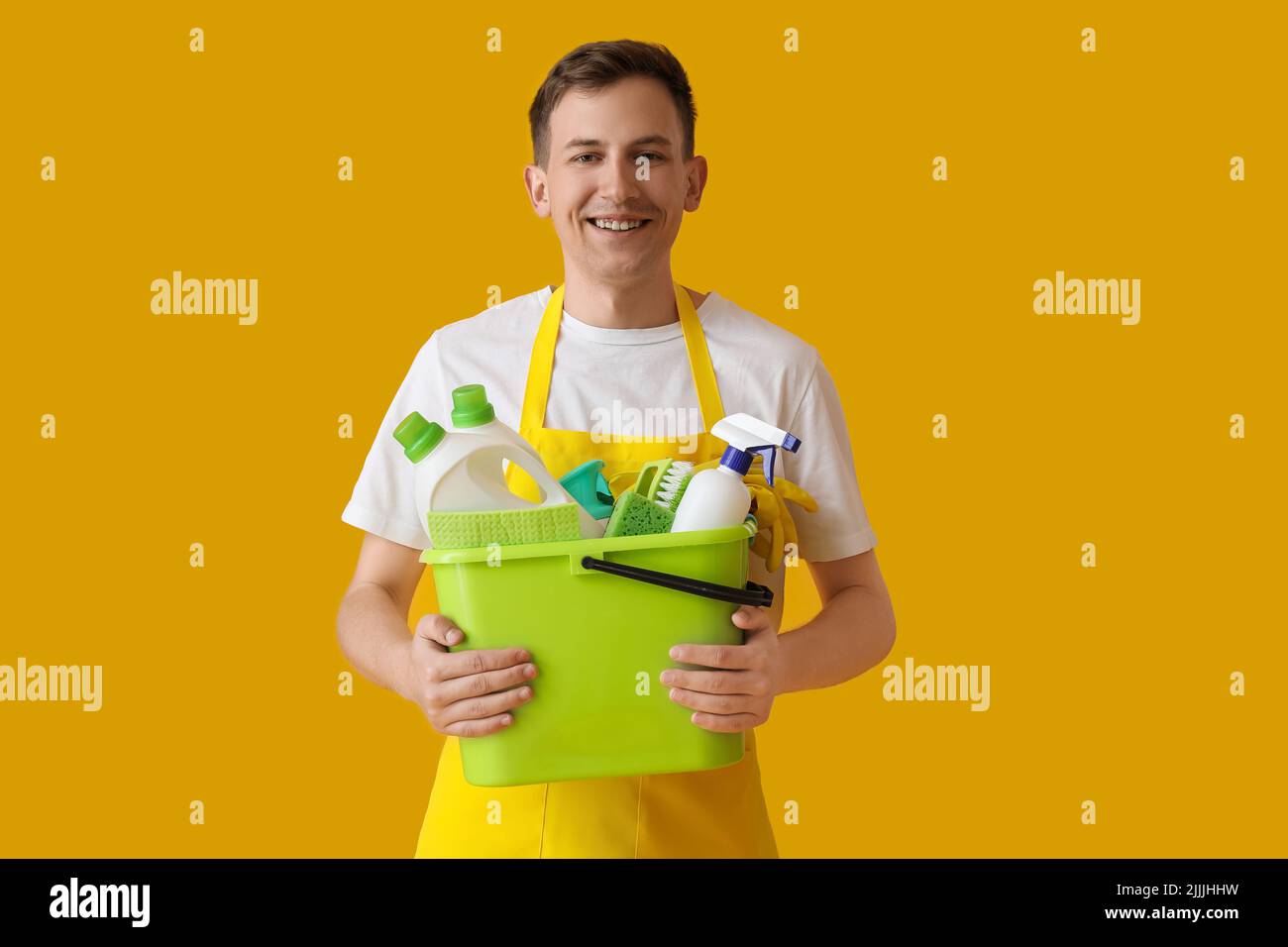 Smiling young man holding bucket with cleaning supplies on yellow ...