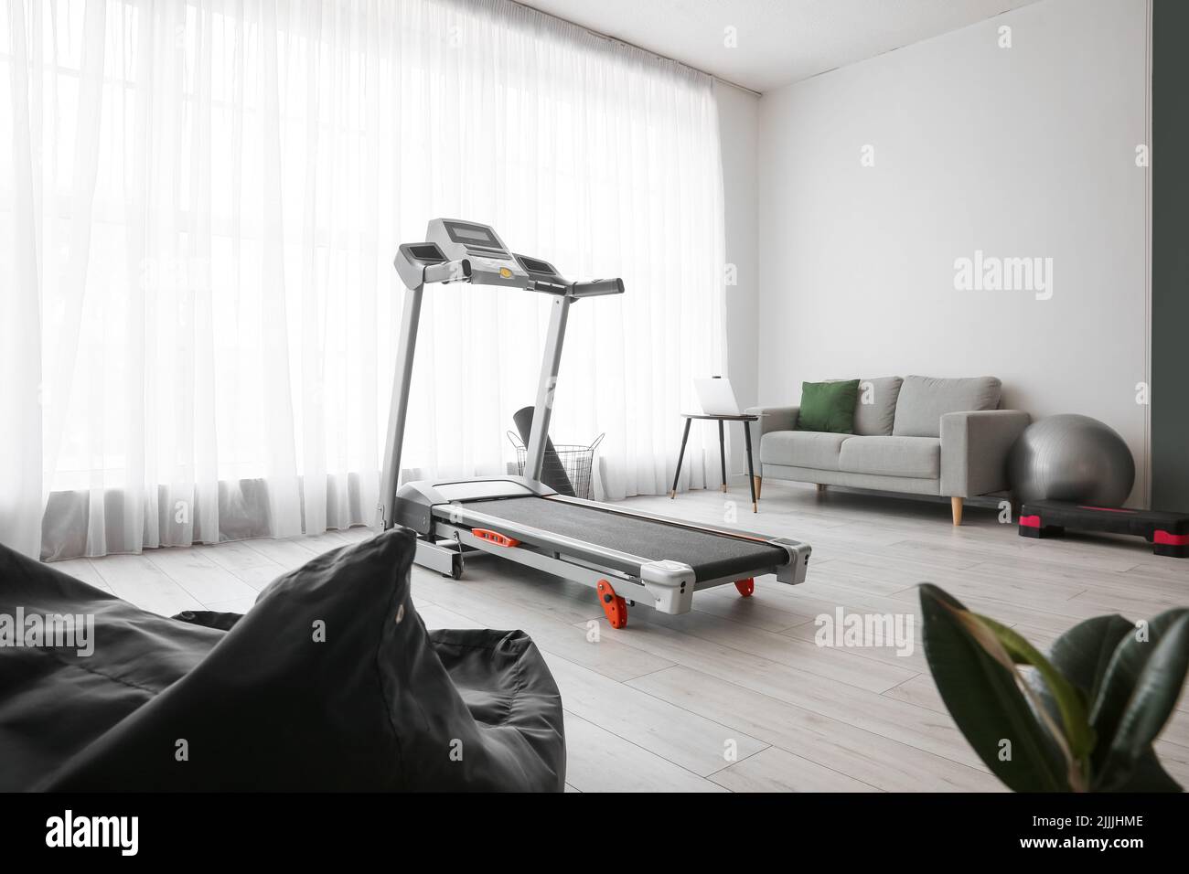 Interior of light living room with treadmill, sofa and sport equipment ...