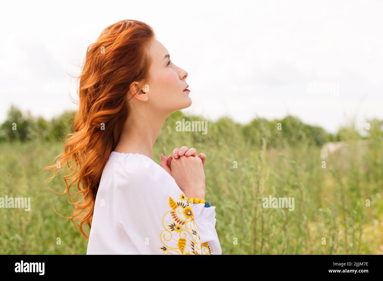 Red-haired, beautiful, young woman in a Ukrainian embroidered shirt prays for Ukraine in the ...