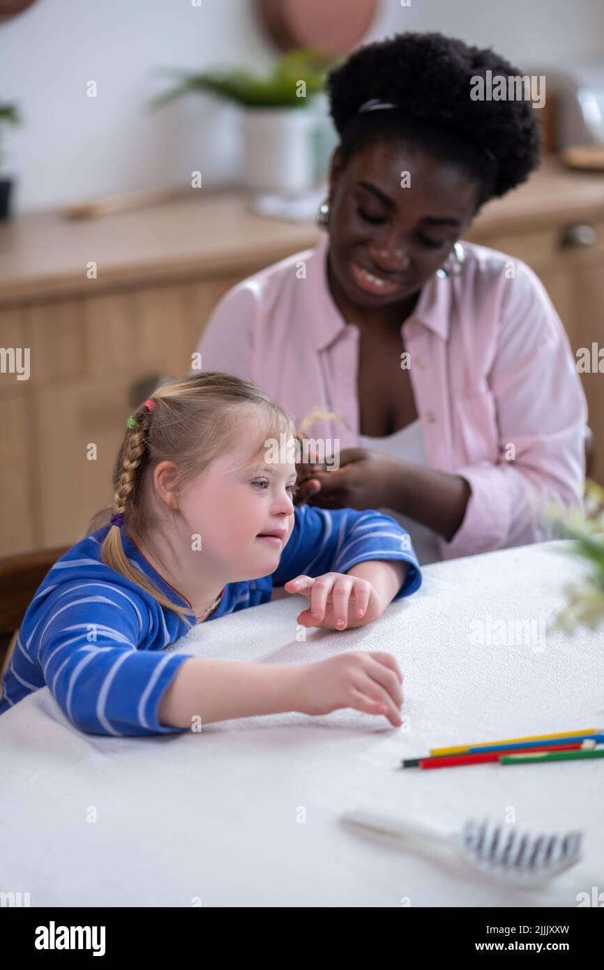 A smiling dark-skinned woman doing hair to the cute girl with down ...