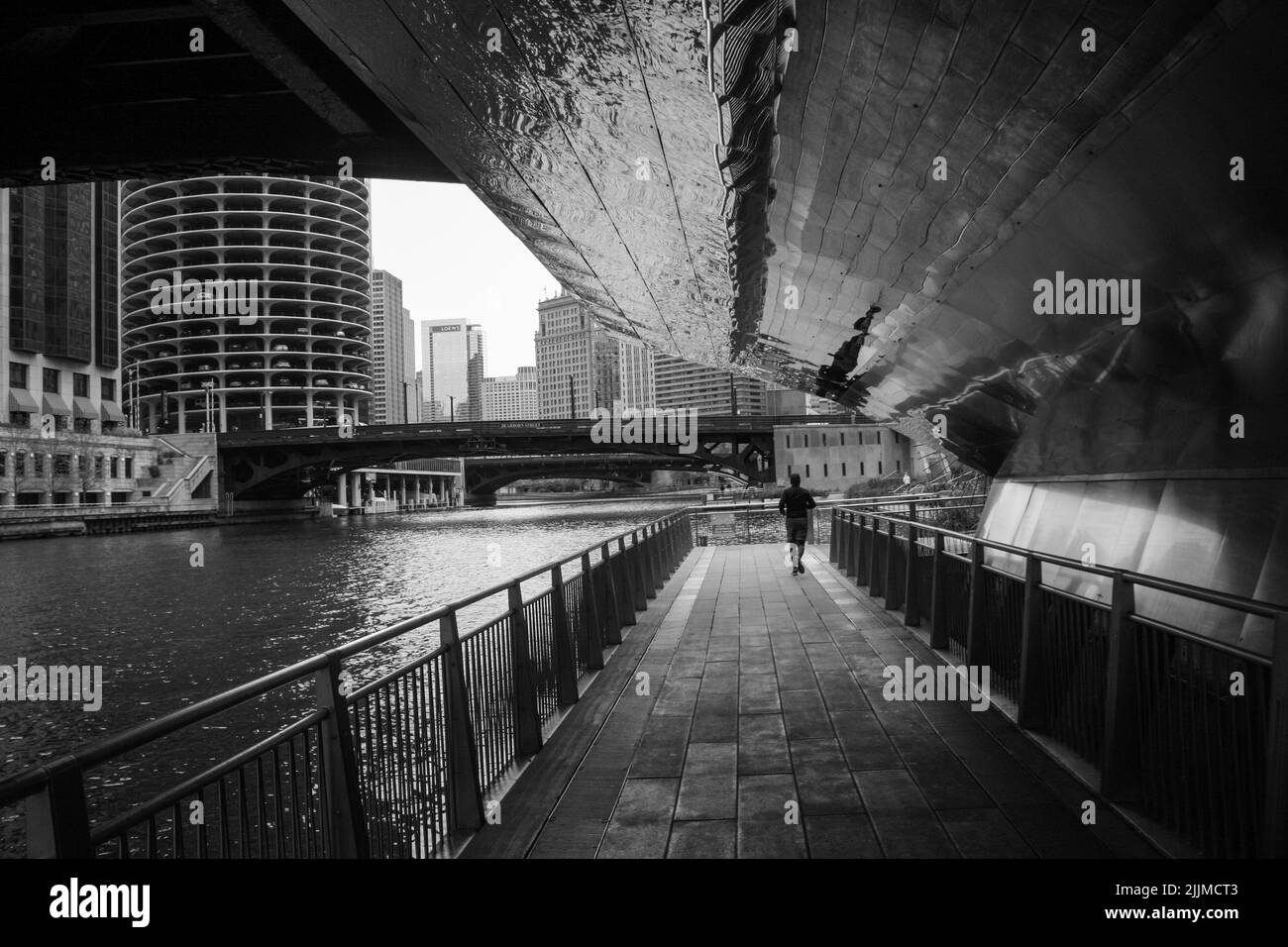 A grayscale of a man running under the bridge near the river Stock ...