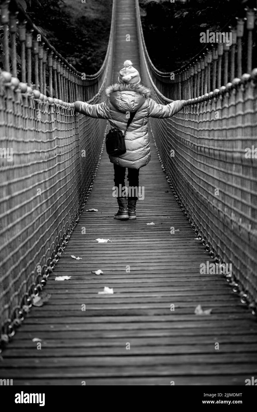 A vertical grayscale shot of a person walking on a suspension bridge ...