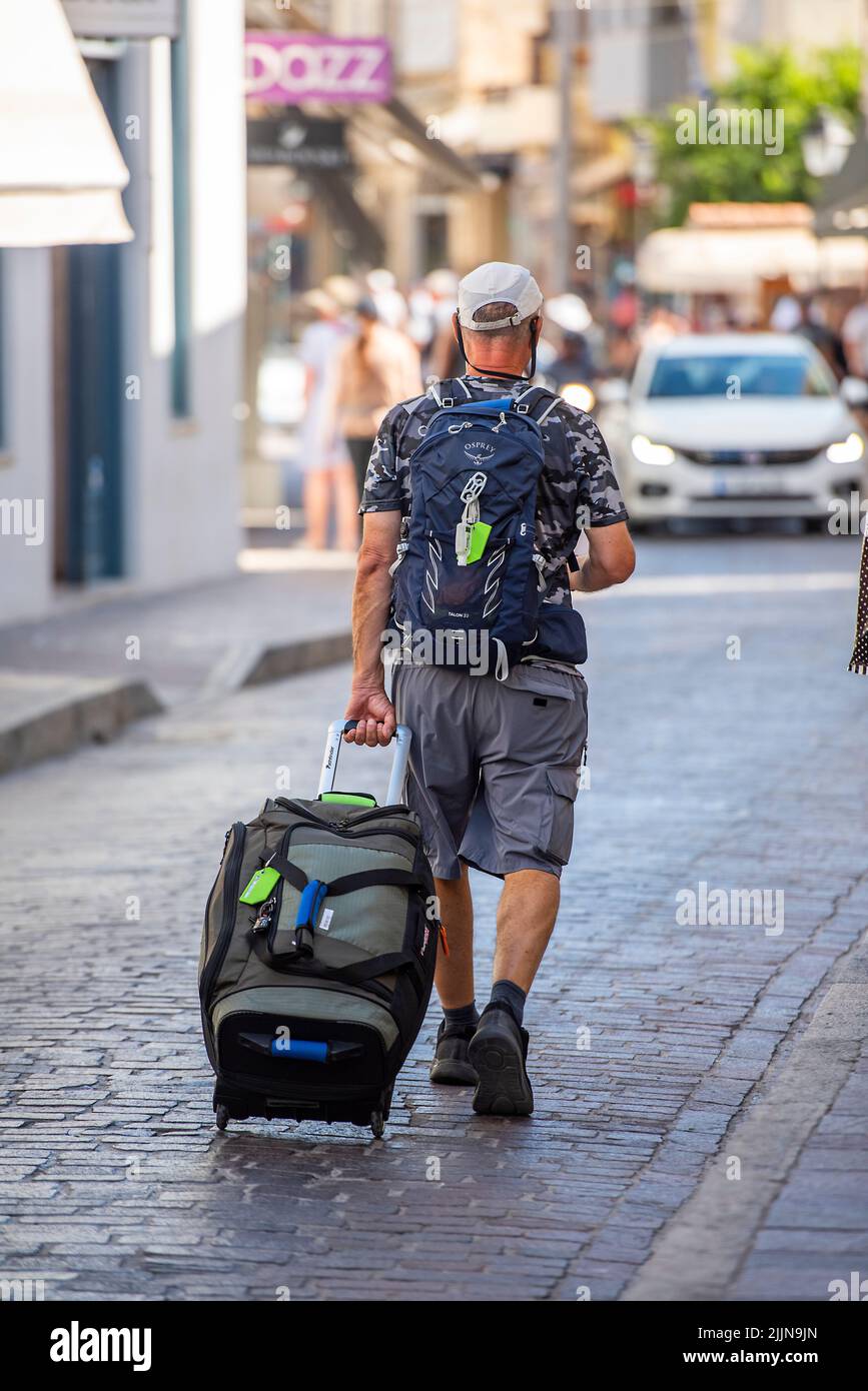 young man walking along street in greece pulling suitcase on wheels ...