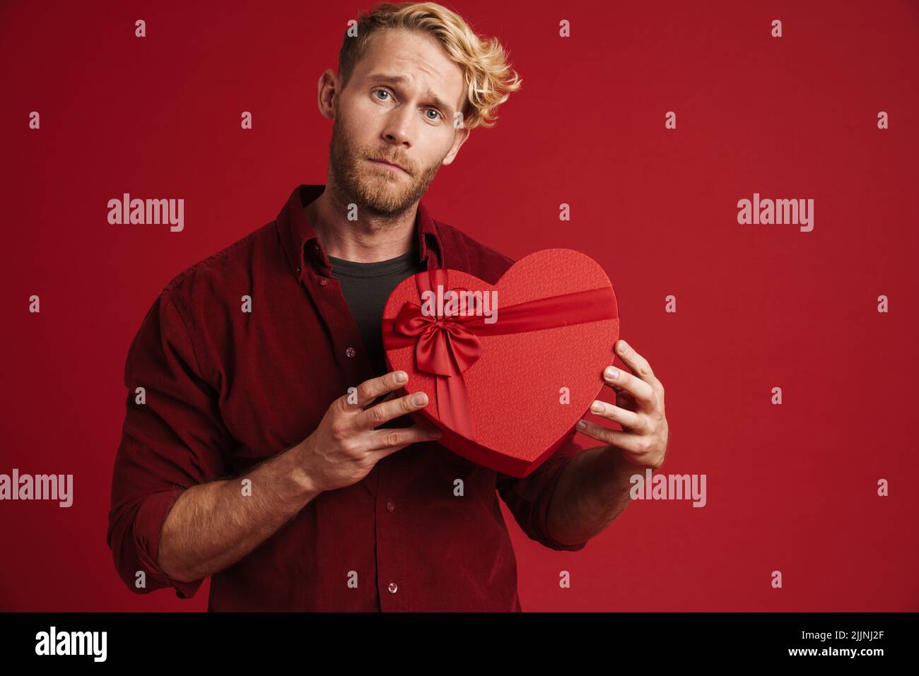 Bearded blonde man frowning while showing heart gift box isolated over ...
