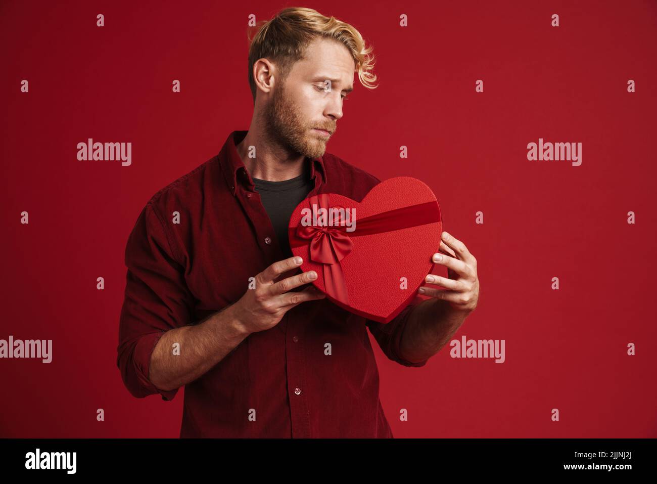 Bearded blonde man frowning while showing heart gift box isolated over ...