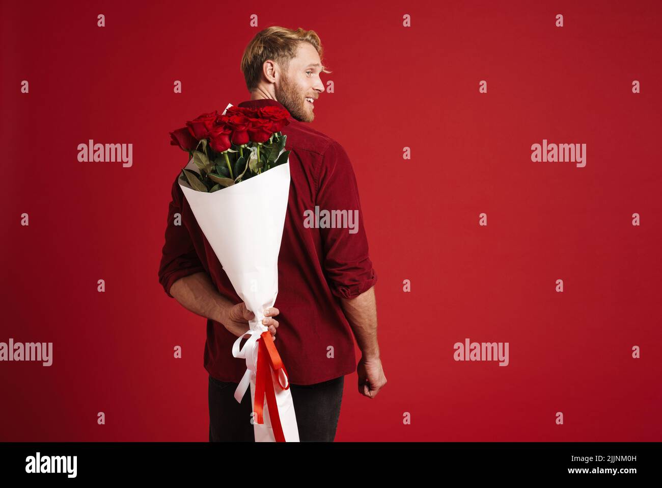 Blonde man smiling while posing with bouquet of roses isolated over red ...