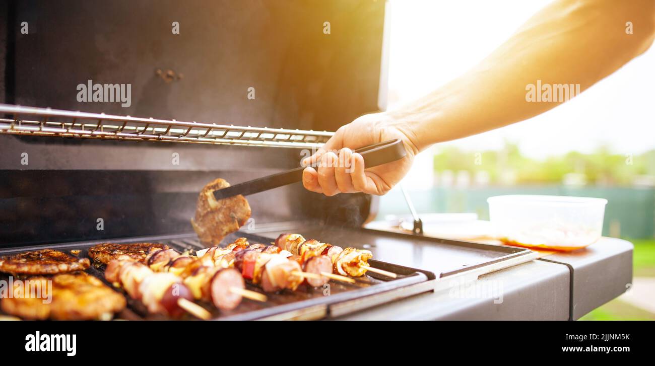 A young man hand is grilling some meat and vegetable skewers on a huge ...