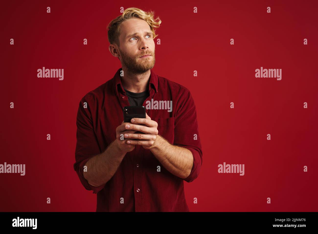 White puzzled man wearing shirt using mobile phone isolated over red ...