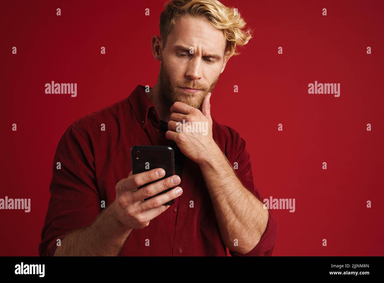 White puzzled man wearing shirt using mobile phone isolated over red ...