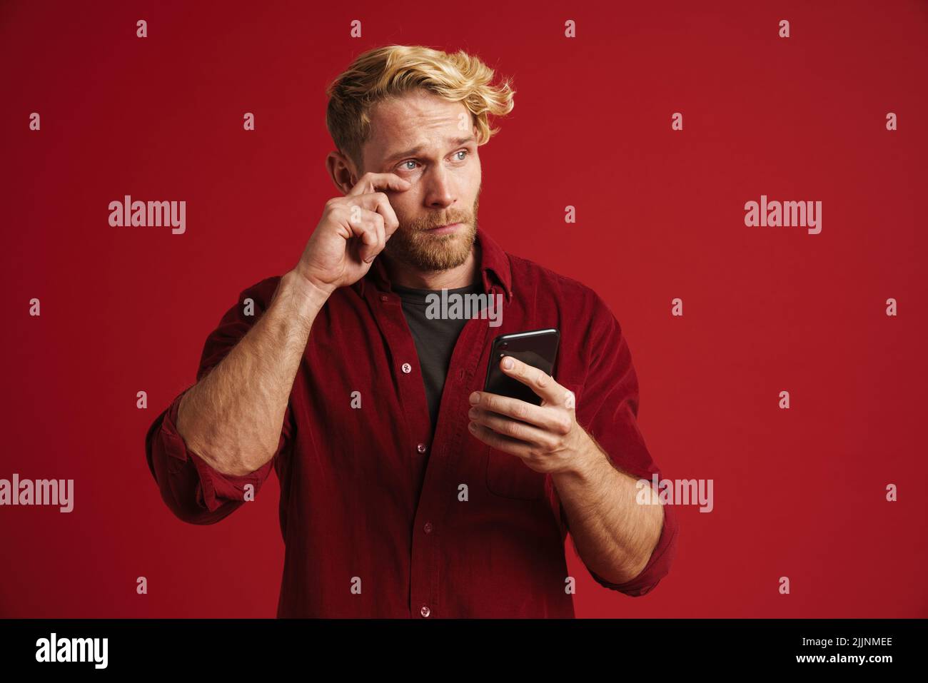 White bearded man crying while using mobile phone isolated over red ...