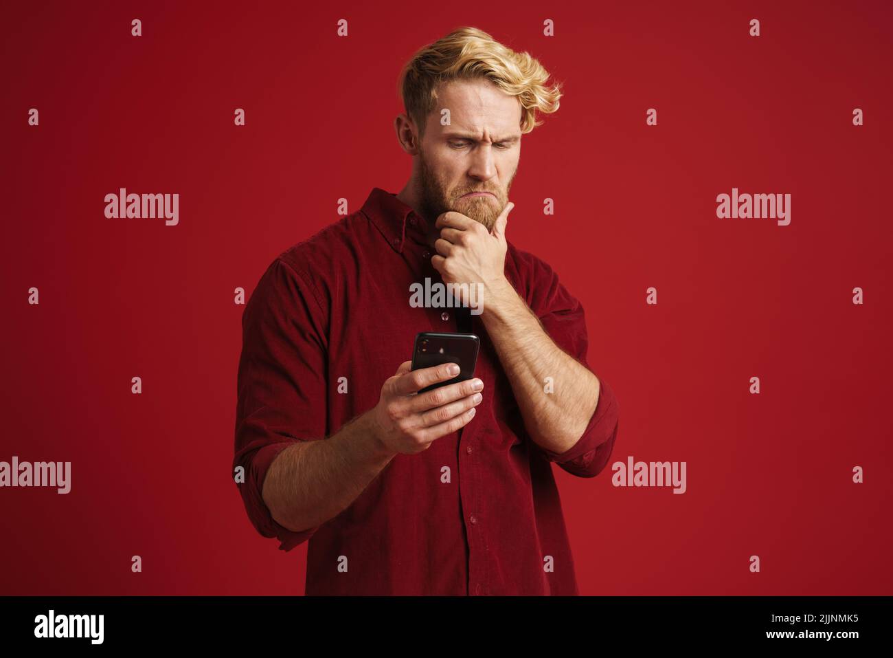 White puzzled man wearing shirt using mobile phone isolated over red ...