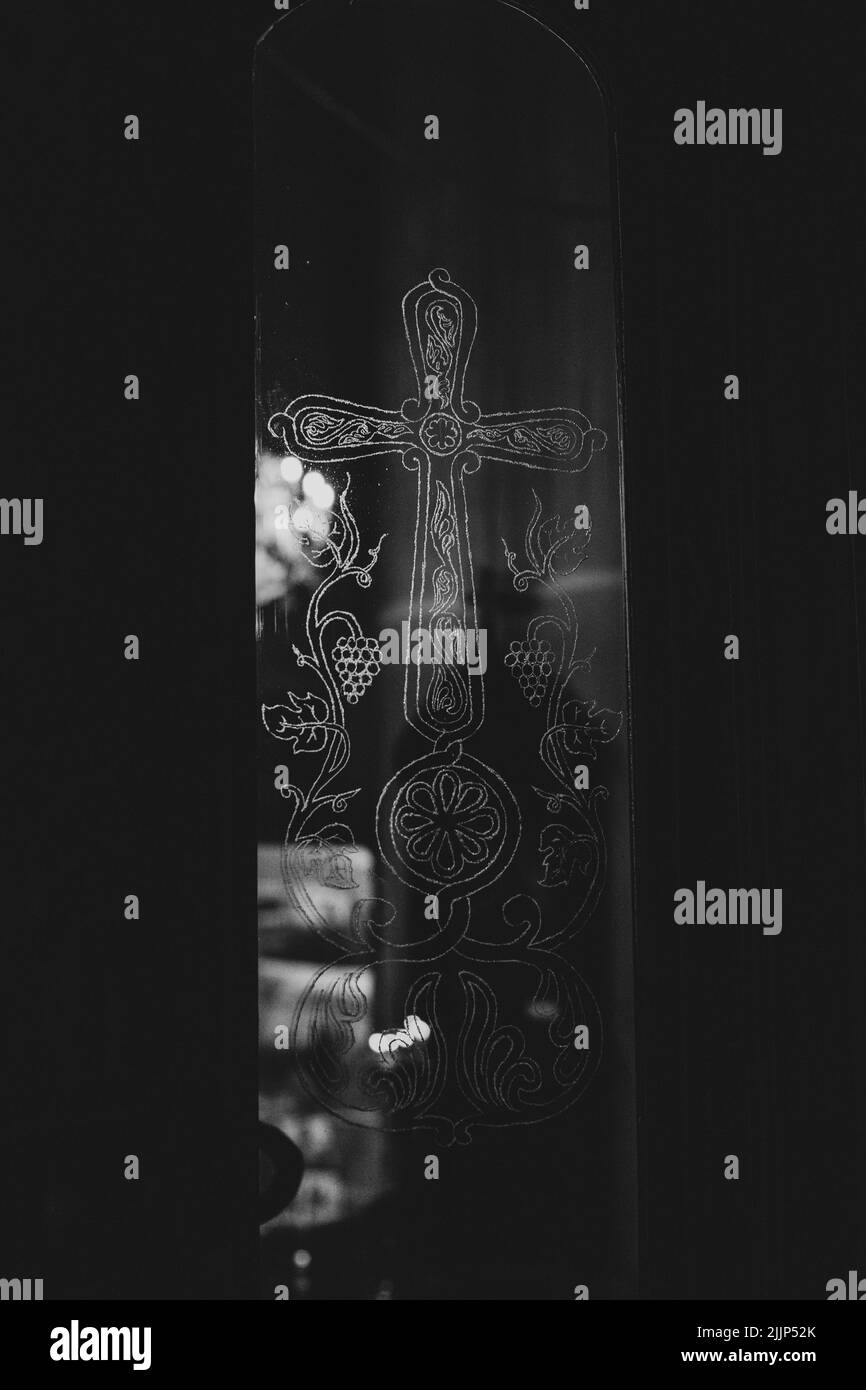 A grayscale vertical shot of a cross drawn on glass of a church door ...