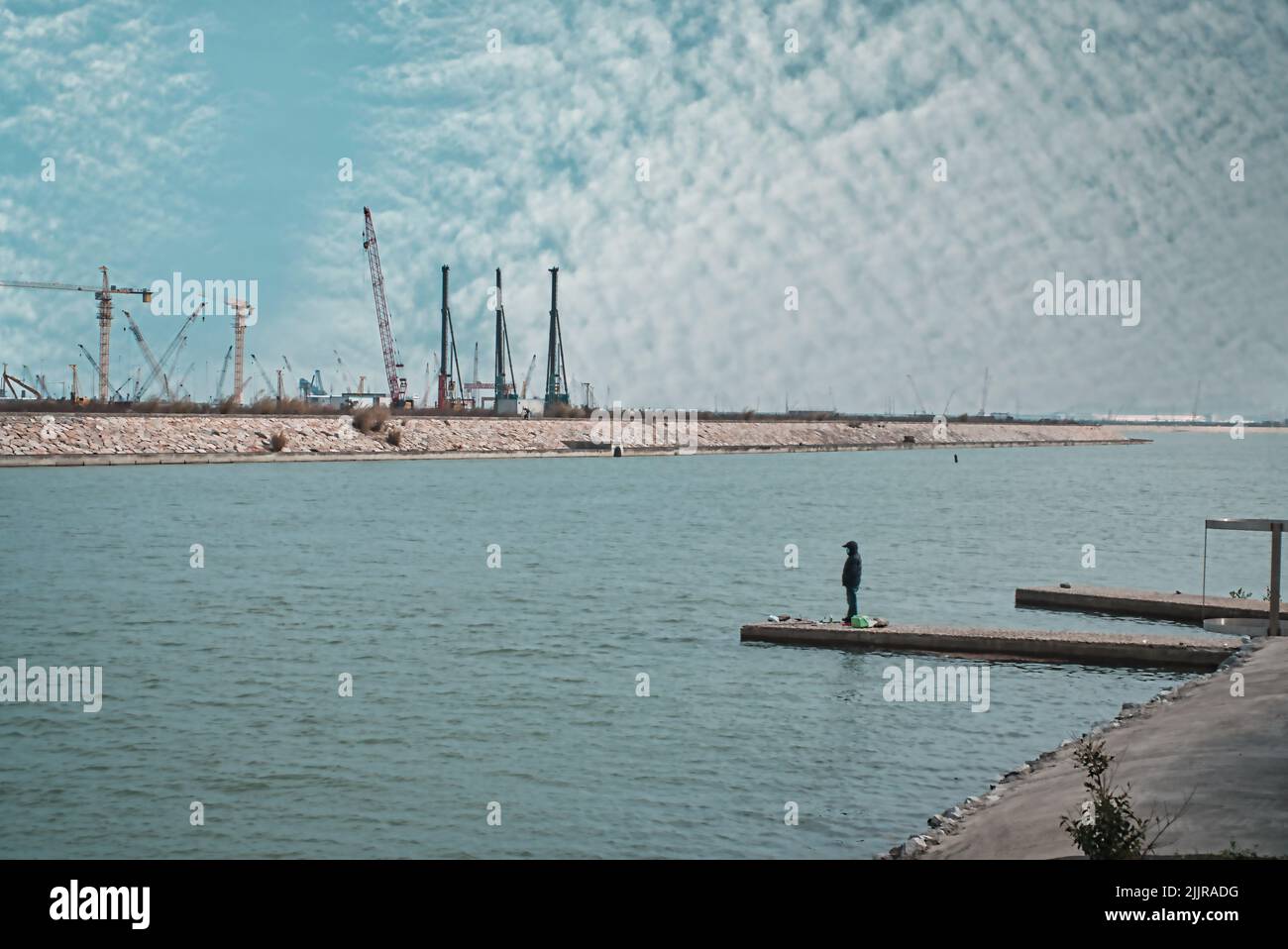 A male fishing near the sea in Macau, China with construction cranes a ...