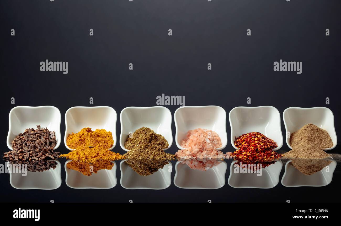 Small white bowls with various spices on a black reflective background ...