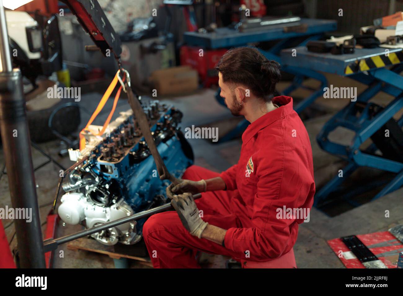 A young Caucasian car mechanic in a red uniform repairing the engine ...