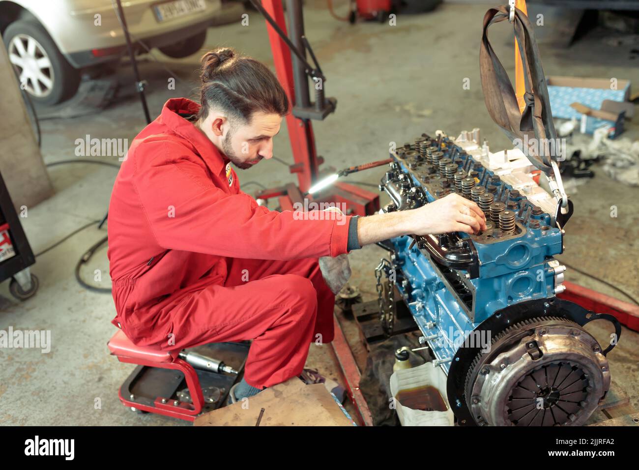 A young Caucasian mechanic in a red uniform mounting the car engine ...
