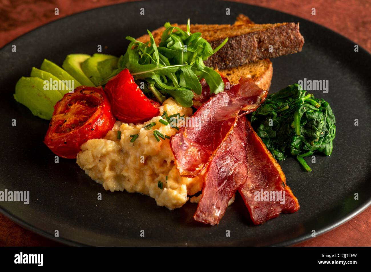 A plate of English breakfast with fried bacon, scrambled eggs, toast, avocado slices and greens ...