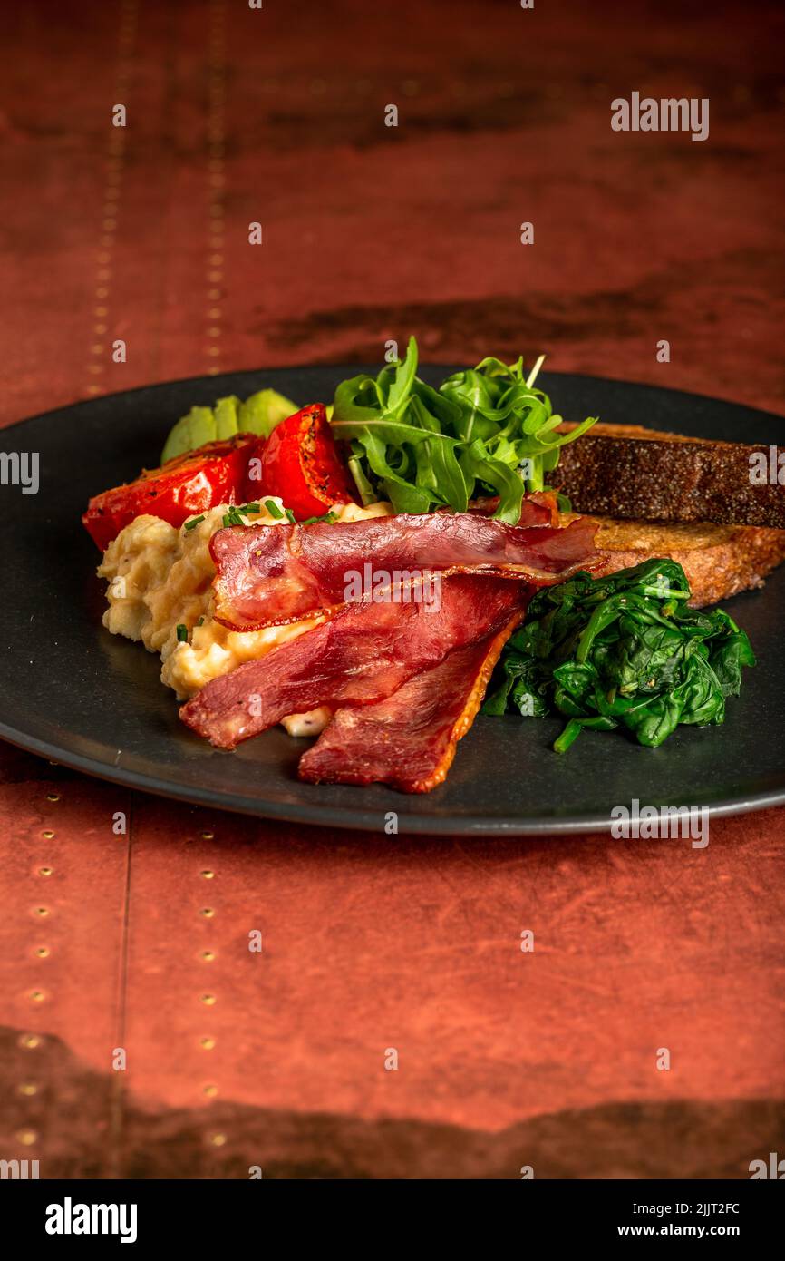 A plate of English breakfast with fried bacon, scrambled eggs, toast, avocado slices and greens ...