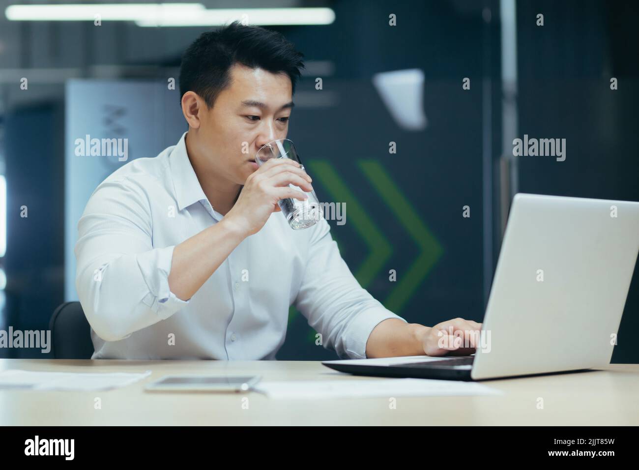 A young Asian businessman drinks water from a glass at a desk in a modern office, works with a ...