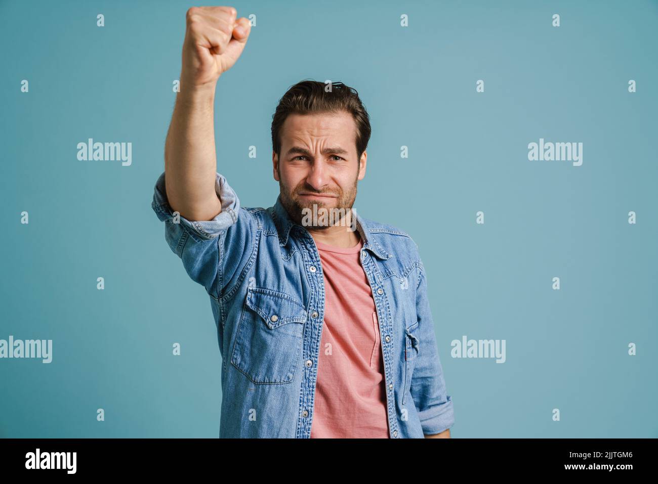Young furious man frowning while shaking his fist isolated over blue ...