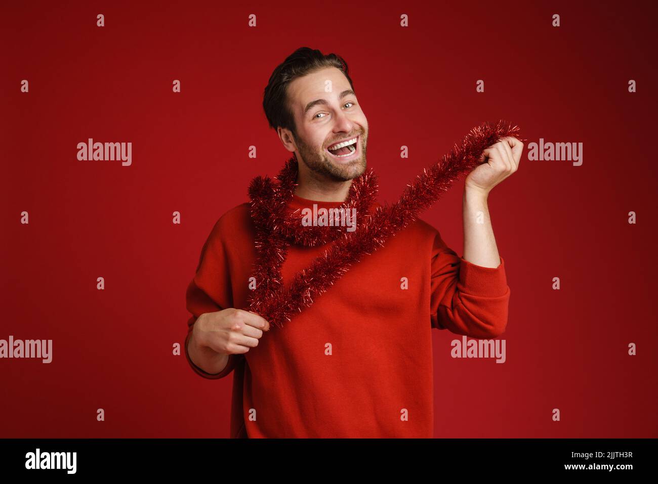 Young bristle man laughing making fun with tinsel isolated over red ...