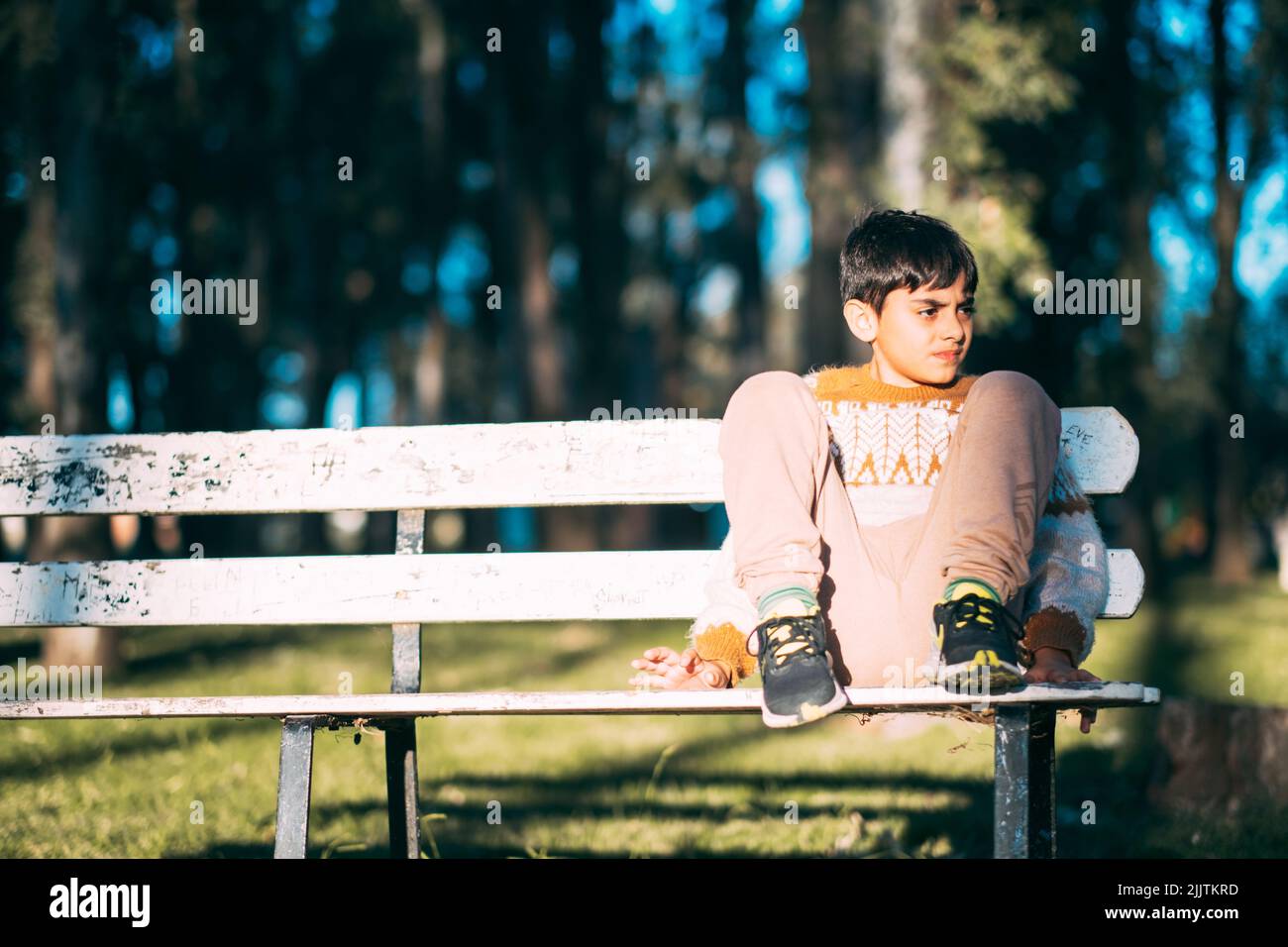 An angry woried Caucasian boy sitting with legs on a bench in a park ...