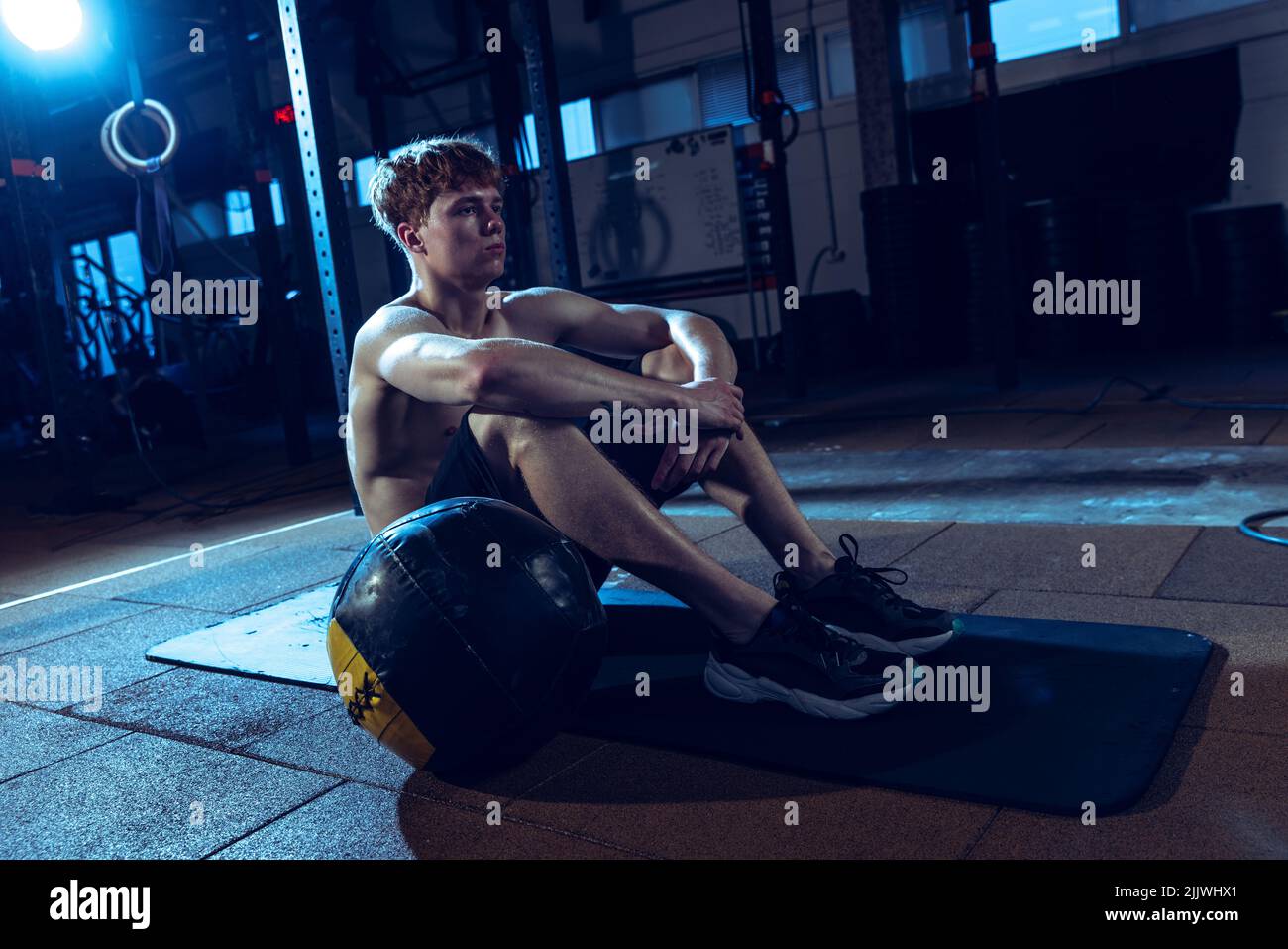 Portrait of young sportive man sitting on fitness rug isolated over gym ...