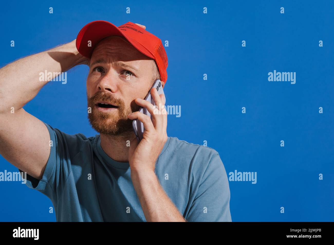 White ginger man wearing cap talking on mobile phone isolated over blue ...