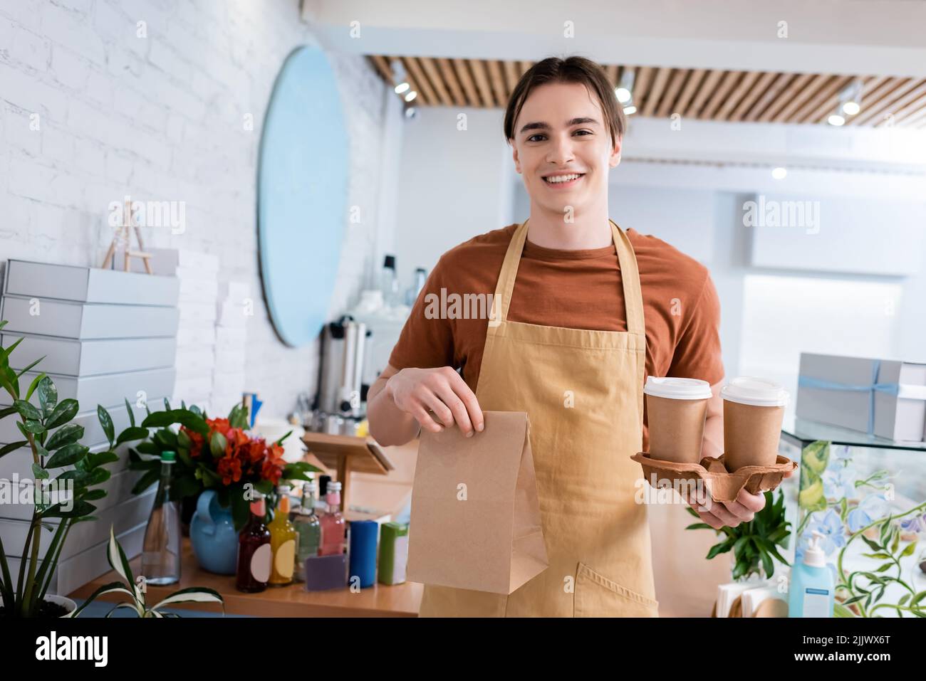 Positive salesman holding coffee to go and paper bag in confectionery ...