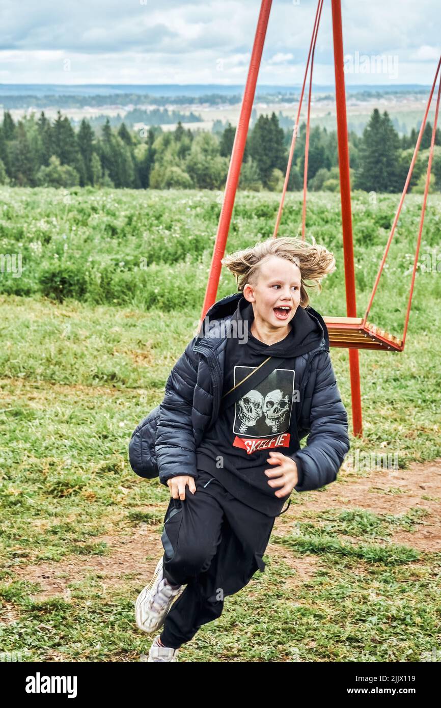 Cheerful boy runs after jumping off swings against lush trees and green ...