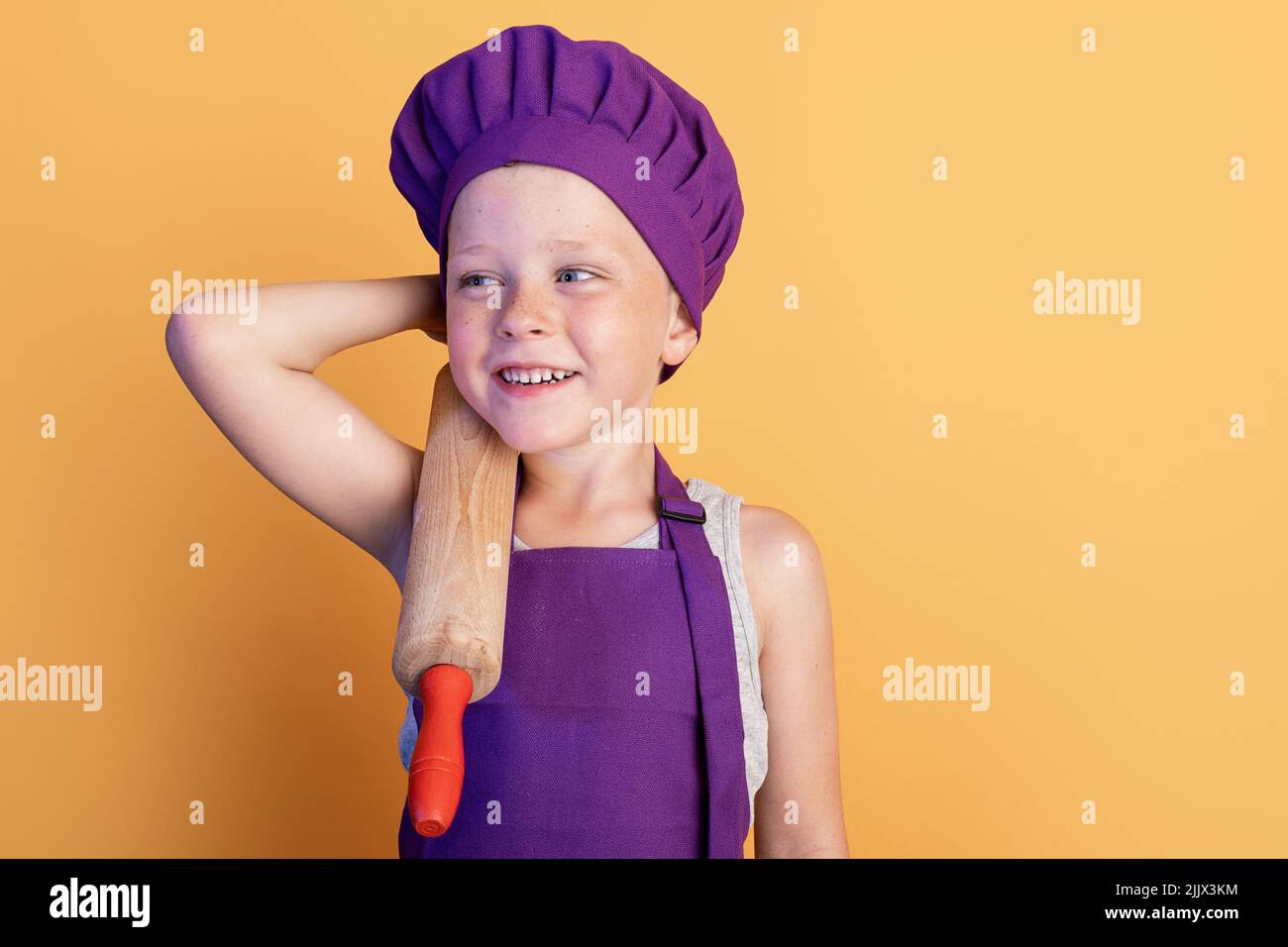 Happy little child in purple chef apron and hat putting rolling pin on ...