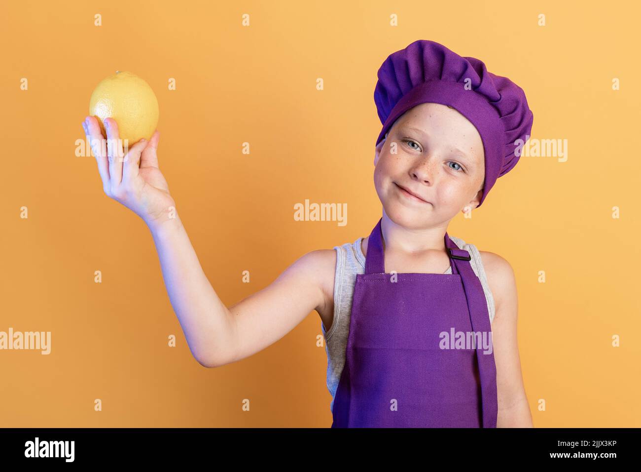 Positive little child in purple chef apron and hat smiling while ...