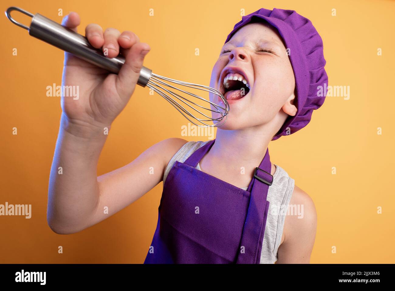 Cheerful boy in purple chef uniform singing into metal whisk with ...