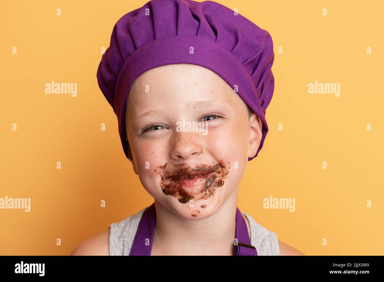 Delighted dirty boy in purple chef hat and apron with face covered with ...